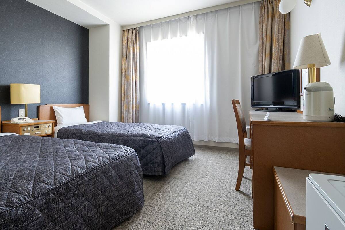 Quarto de hotel com duas camas, uma escrivaninha com televisão, uma lâmpada e uma cafeteira.