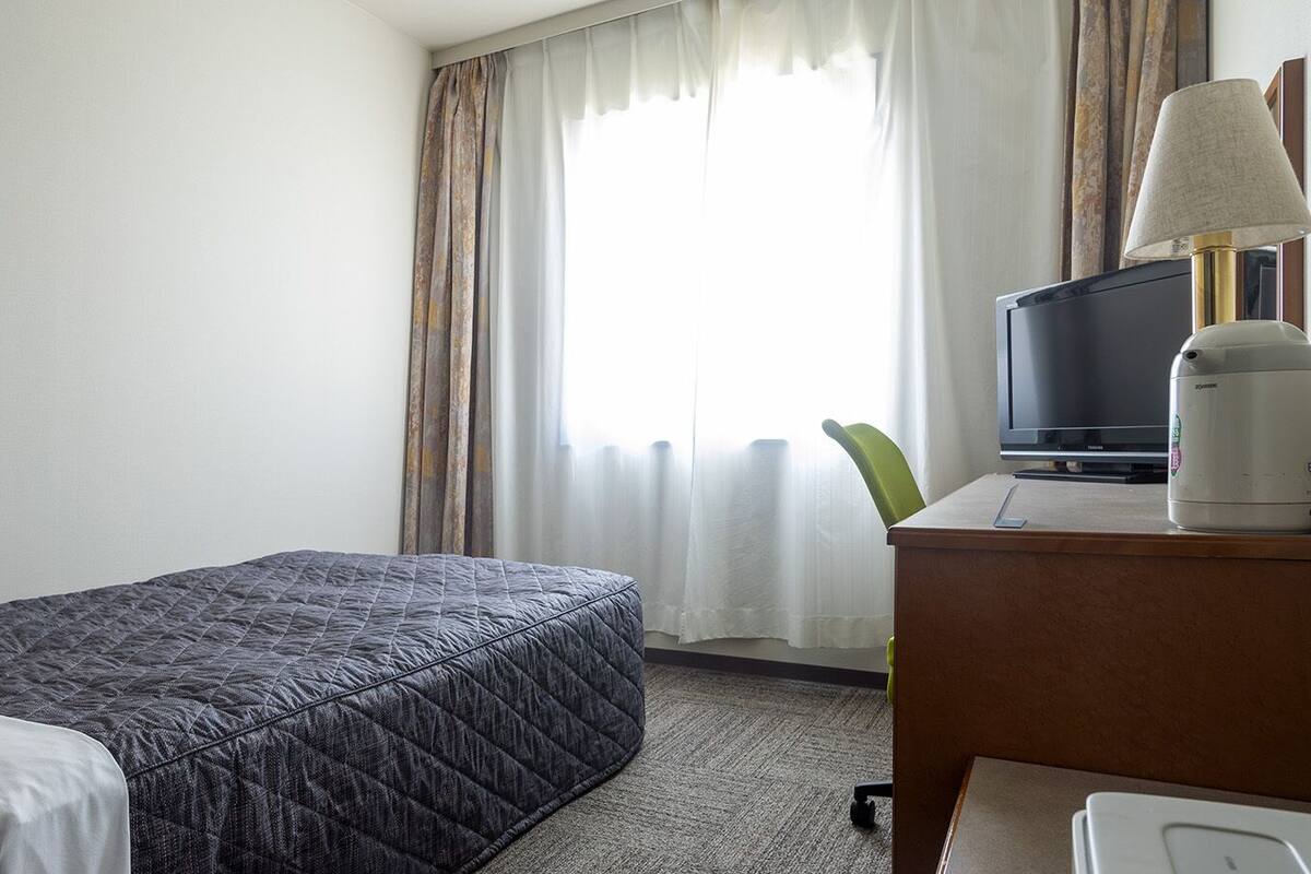 Quarto de hotel com cama, televisão, luminária e escrivaninha.