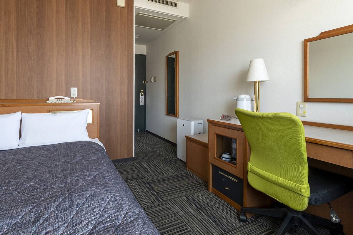 Quarto de hotel com cama, escrivaninha, cadeira, espelho, abajur e pequena cozinha.
