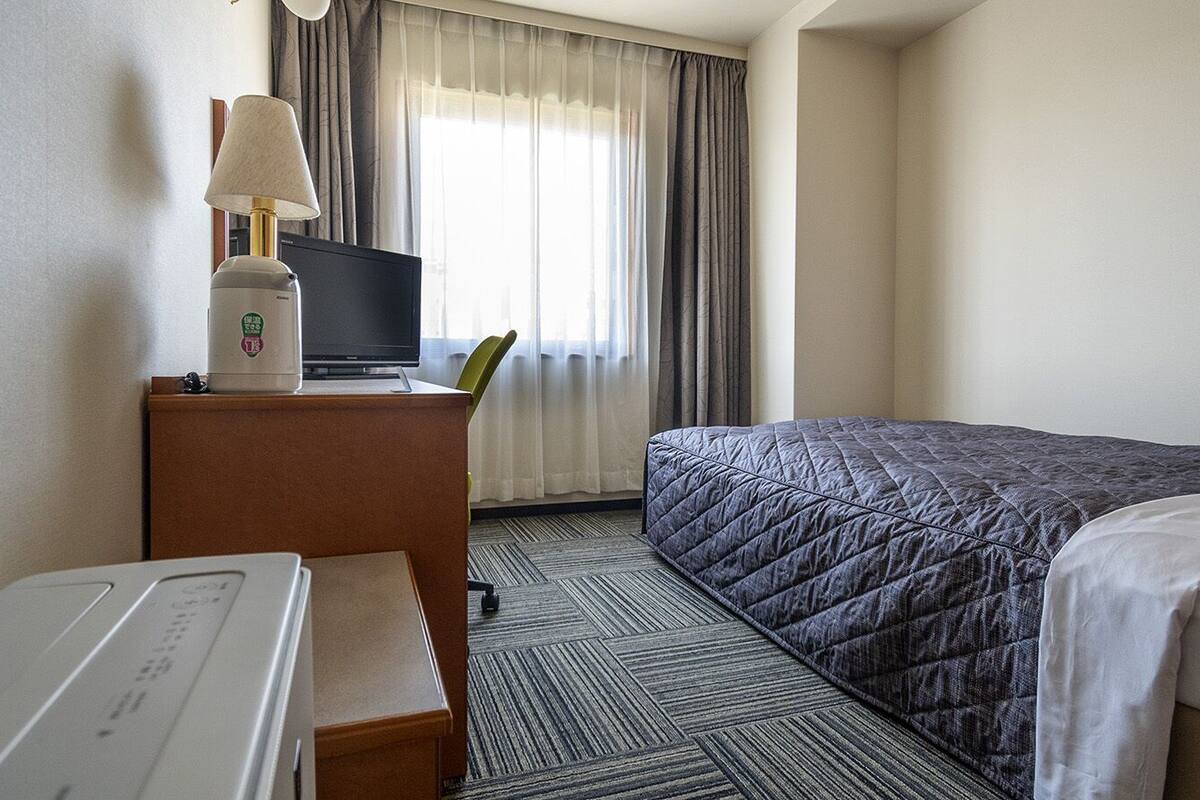 Quarto de hotel com cama, escrivaninha com luminária, televisão e janela com cortinas.