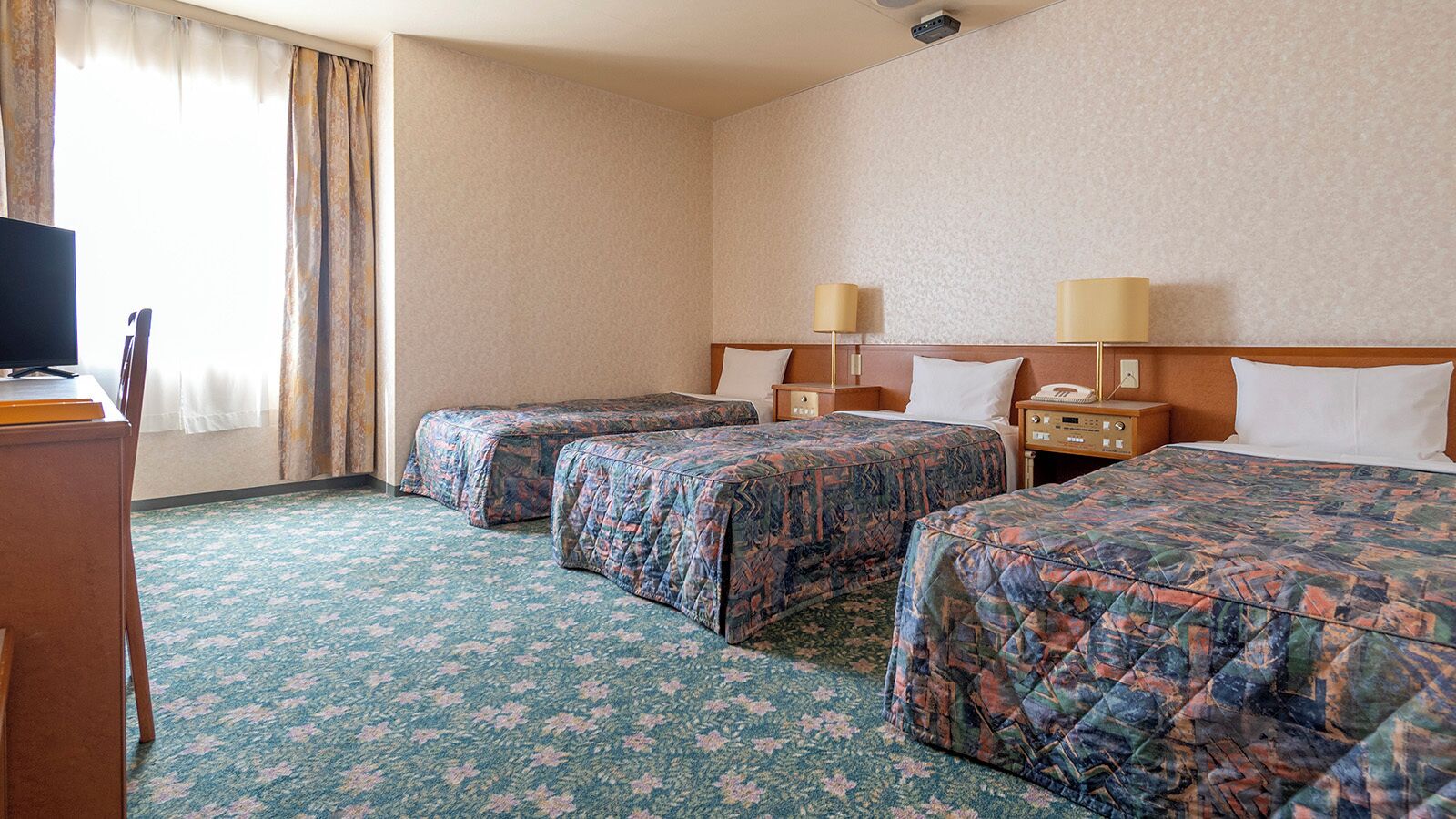 Quarto de hotel com três camas, uma escrivaninha e uma televisão.