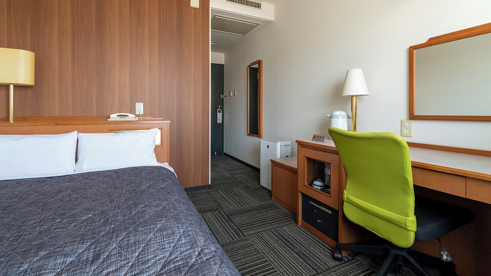 Quarto de hotel com cama, escrivaninha, cadeira, espelho, luminária e pequeno armário.
