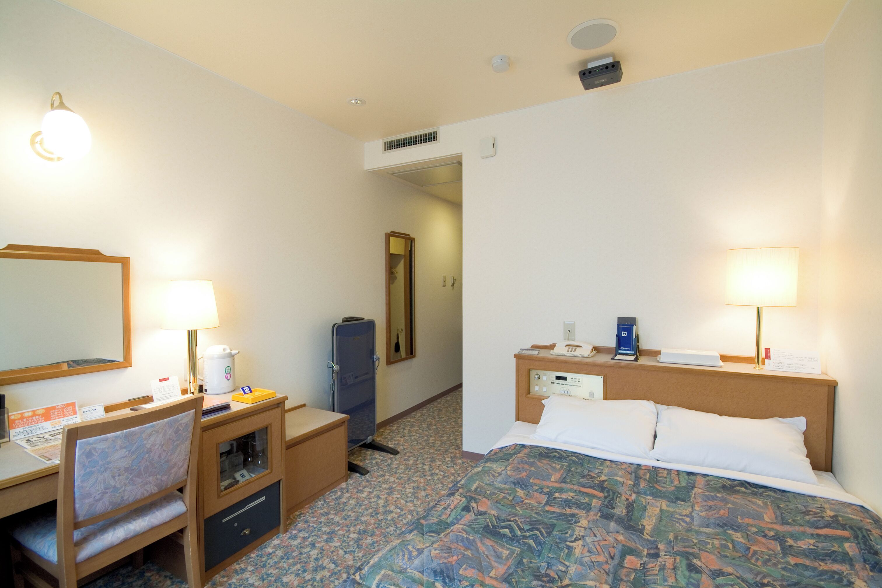 Quarto de hotel com cama, escrivaninha, cadeira e espelho.