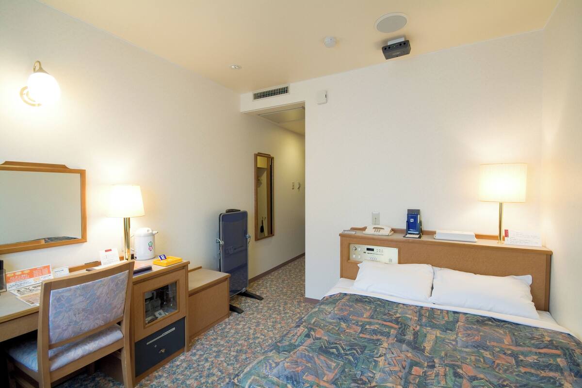 Quarto de hotel com cama, escrivaninha, cadeira e espelho.