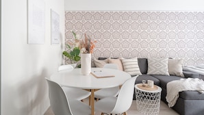 Uma sala de estar moderna com uma mesa de jantar redonda, cadeiras brancas, um sofá cinza e papel de parede estampado.