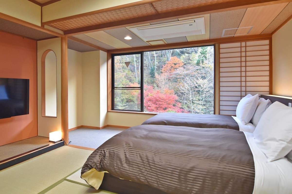 Quarto de hotel com piso de tatame, cama com roupa de cama branca, televisão na parede e uma grande janela com vista para a folhagem de outono.