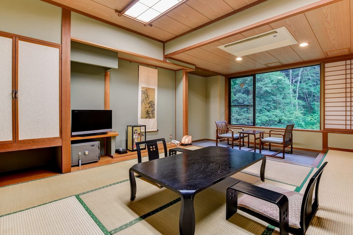 Quarto em estilo japonês tradicional, com piso de tatame, uma mesa baixa, cadeiras, televisão e uma janela grande com vista para uma floresta.