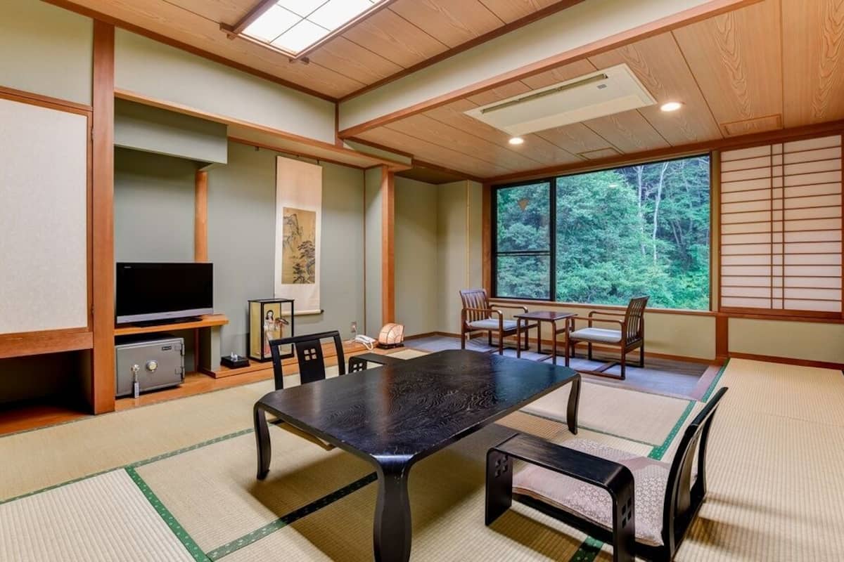 Um quarto japonês tradicional com tatames, uma mesa baixa, cadeiras, televisão e uma porta deslizante com vista para uma floresta.
