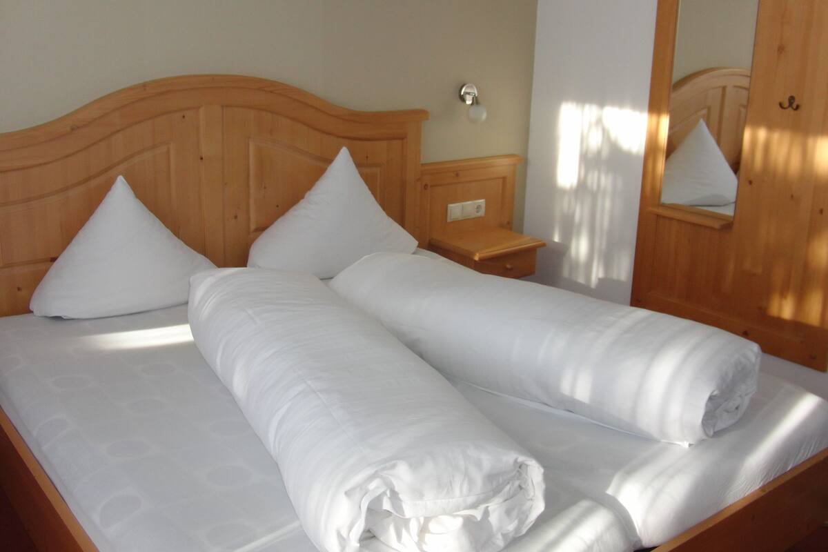 Uma cama de casal com cabeceira de madeira e almofadas, um criado-mudo de madeira e um espelho na parede.