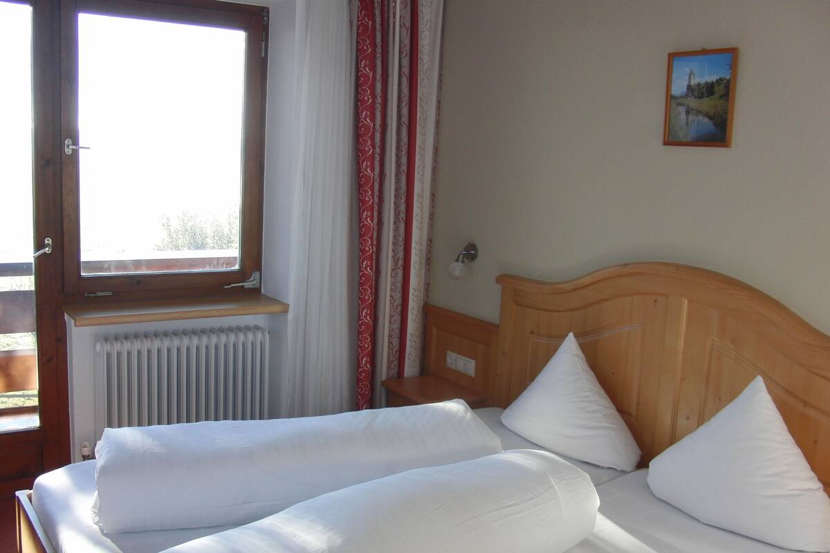 Quarto com cama de madeira, roupa de cama branca, janela com cortinas e um quadro na parede.