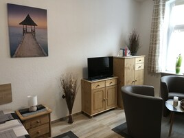 Uma sala de estar com televisão sobre um aparador de madeira, um sofá, uma mesa pequena com uma vela, um vaso com plantas secas e um quadro de um cais ao pôr do sol.