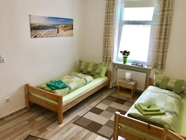 Um quarto pequeno e arrumado com duas camas de madeira, uma janela com cortinas, um radiador e uma pequena mesa.