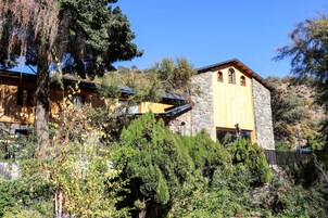 Um edifício de pedra com detalhes em madeira e janelas em arco, rodeado por vegetação densa e árvores.