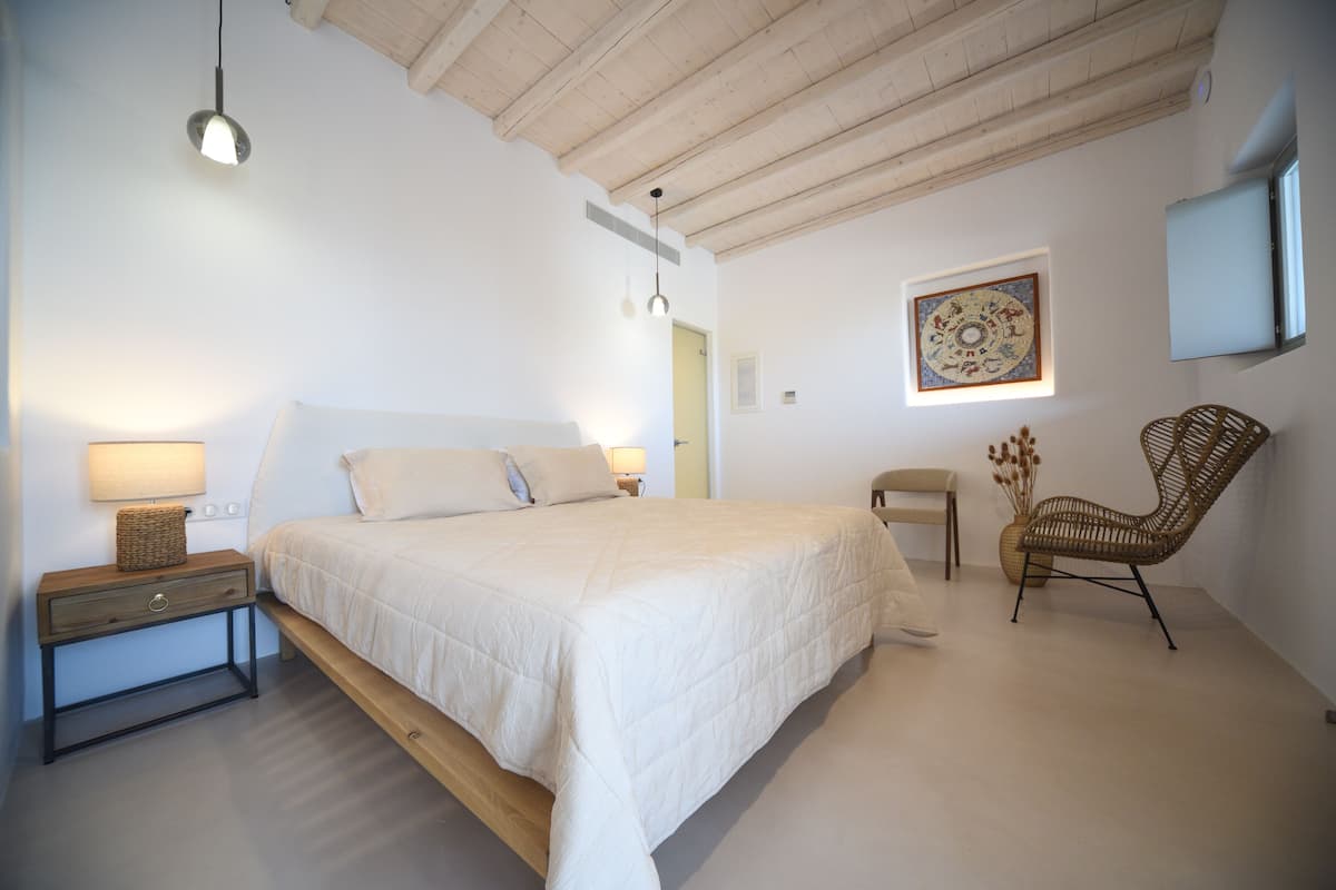 Quarto com cama, abajures, uma cadeira, TV fixada na parede e um objeto decorativo na parede.