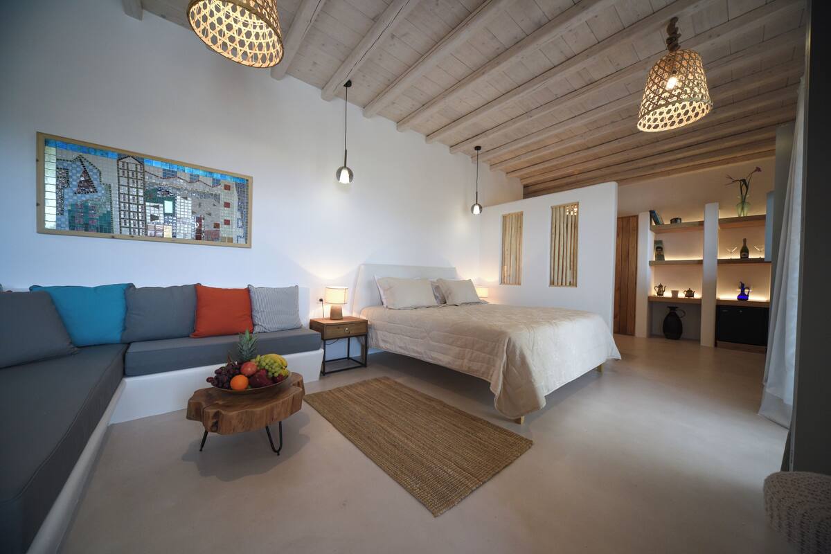 Quarto moderno com uma cama grande, um sofá e uma pequena mesa com frutas. Há luminárias pendentes e uma obra de arte com vista para a cidade na parede.