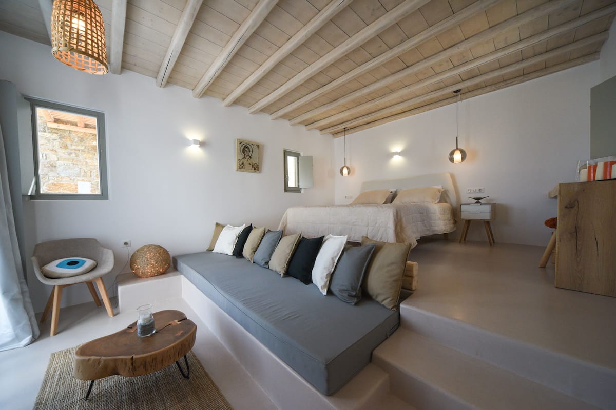 Quarto moderno e bem iluminado com um sofá cinza grande, uma mesa de centro de madeira e uma pequena mesa lateral. Há uma janela com vista para uma parede de pedra, e um teto de madeira com vigas aparentes.