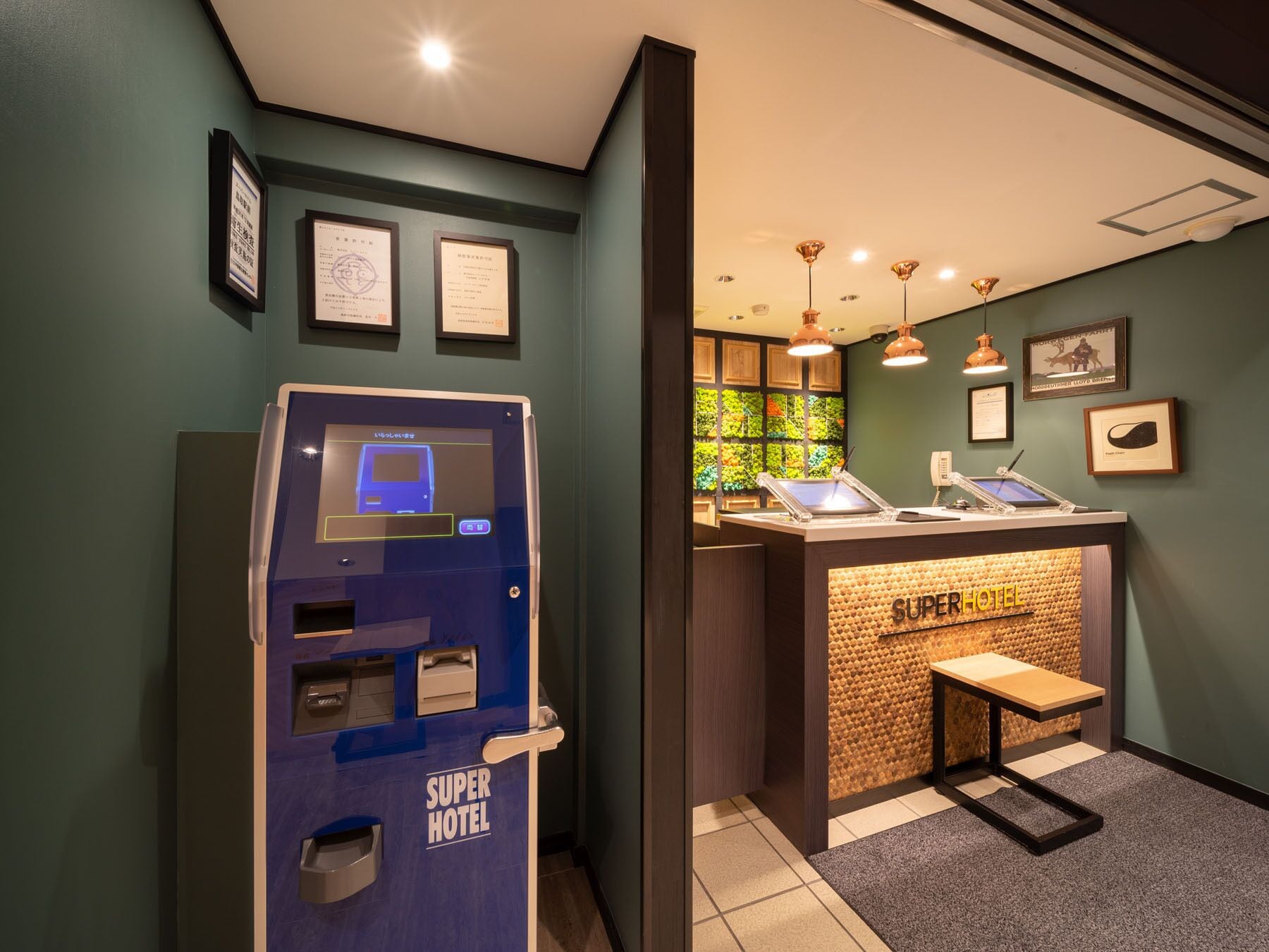 Uma área de recepção com um quiosque azul de autoatendimento identificado como "Super Hotel" e um balcão de recepção com uma placa "Super Hotel".