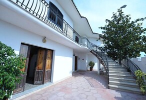 Um edifício branco com porta de madeira, uma varanda com grades pretas e uma escadaria que leva a um andar superior.