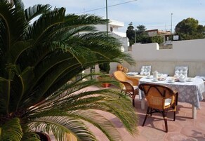 Um pátio externo com mesa posta para refeições, rodeado por cadeiras de vime e uma grande palmeira em primeiro plano.