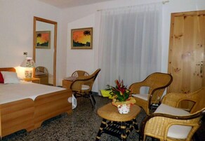 Quarto de hotel com uma cama, duas cadeiras de vime, uma mesinha com um vaso, uma porta de madeira e duas pinturas na parede.