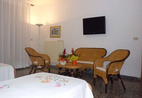 Quarto de hotel com cama, duas cadeiras de vime, uma mesinha com um vaso de flores, televisão de tela plana na parede e um quadro.