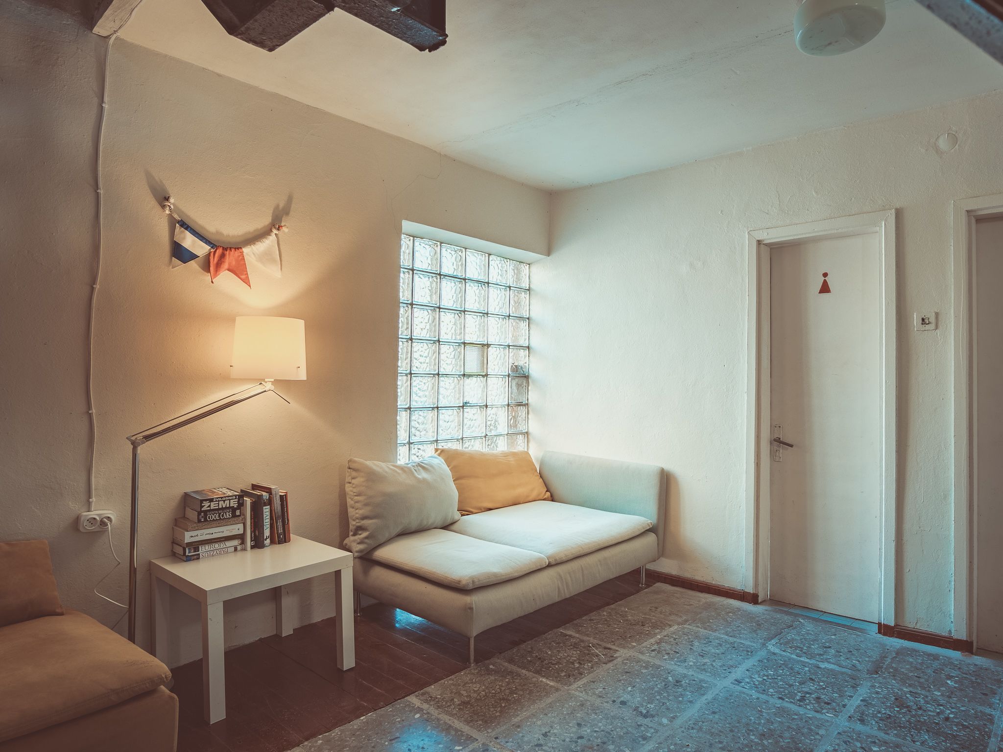 Uma sala de estar aconchegante com um sofá branco, uma mesa pequena com livros, uma luminária e uma janela com um objeto decorativo acima.