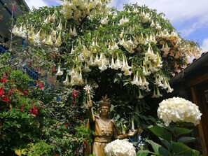 Uma estátua segura um bastão com uma grande flor branca em uma mão e uma flor branca menor na outra, diante de um jardim exuberante com flores brancas e folhagem verde.