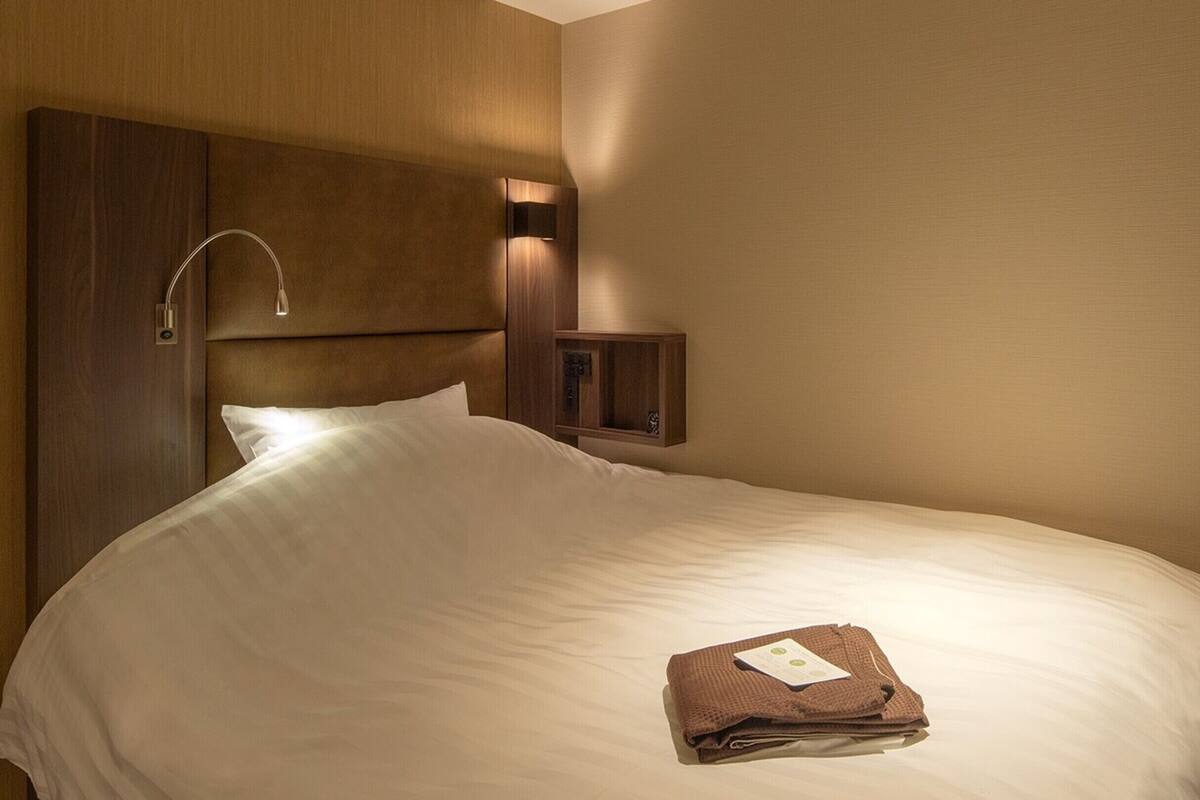 Uma cama bem arrumada com lençóis brancos, uma toalha marrom dobrada e uma pequena mesa de cabeceira de madeira com um abajur.