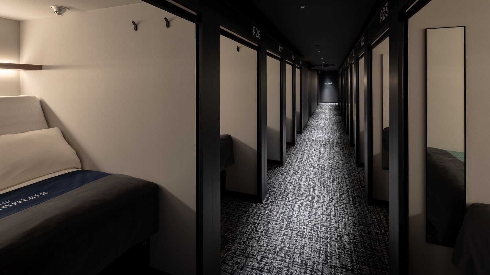 Corredor de hotel com piso de carpete, cama com colcha azul e uma fileira de portas à direita.