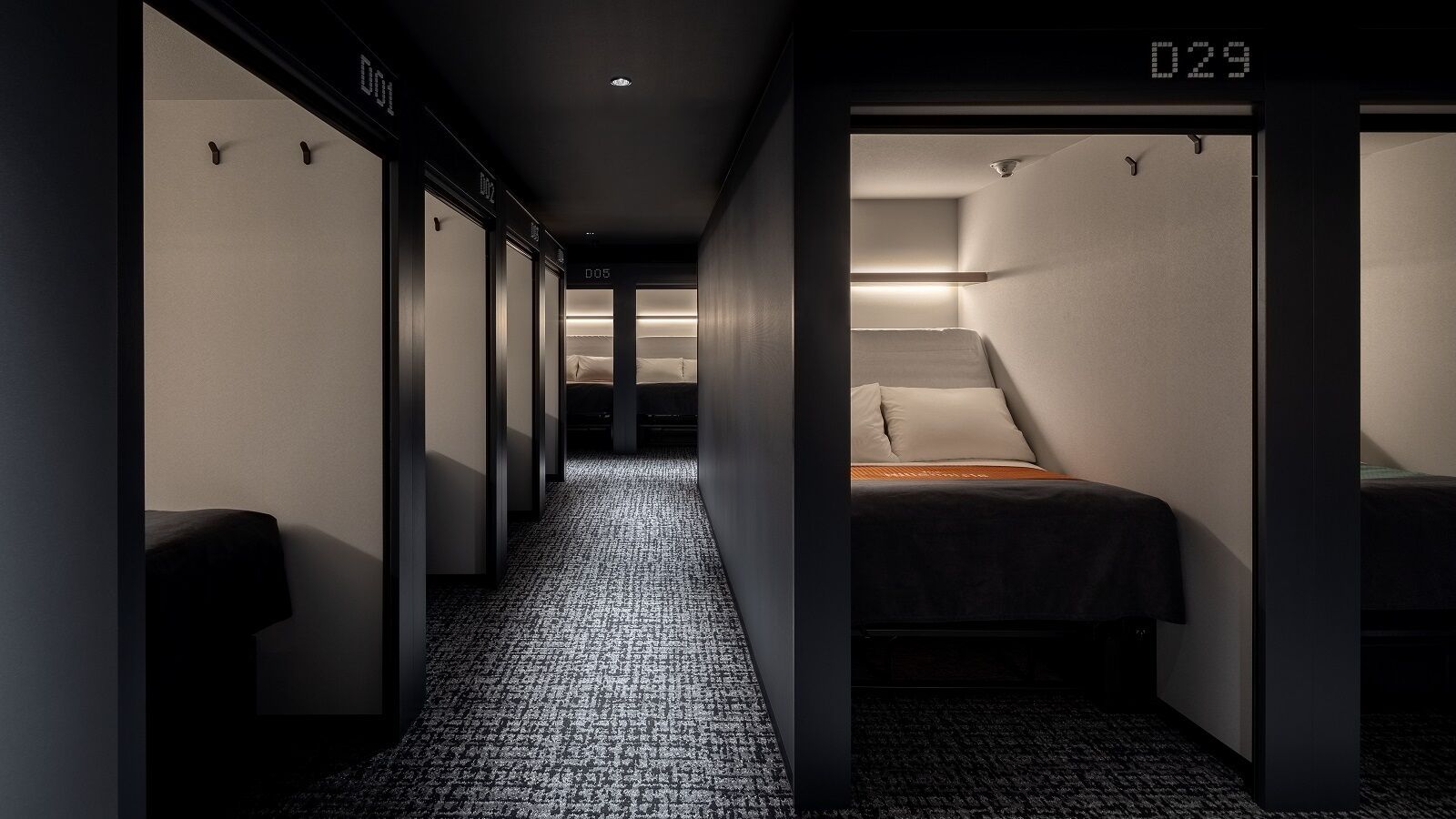 Um corredor estreito com carpete estampado, uma cama com colchão e almofadas, e um relógio digital mostrando as horas.