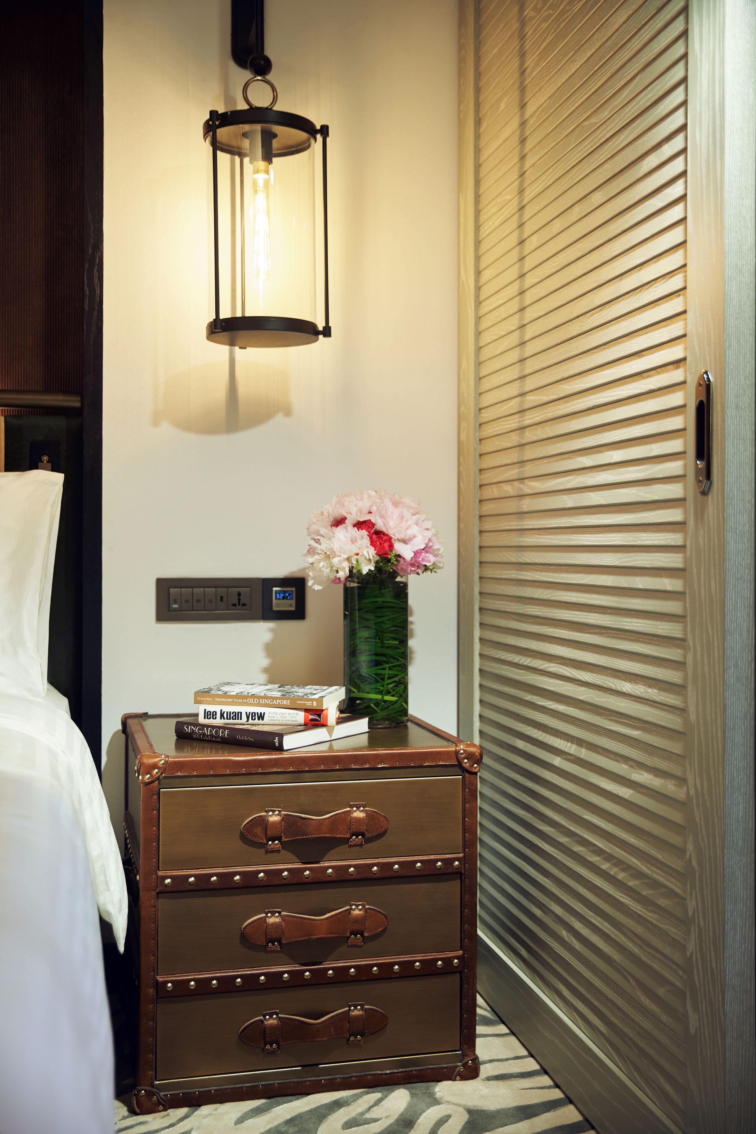 Quarto de hotel com cama, mesa de cabeceira com abajur, vaso com flores e cômoda de madeira.