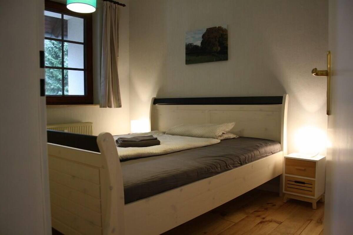 Uma cama de solteiro com cabeceira, uma mesinha de cabeceira com abajur, uma janela com cortinas e um quadro pendurado na parede.