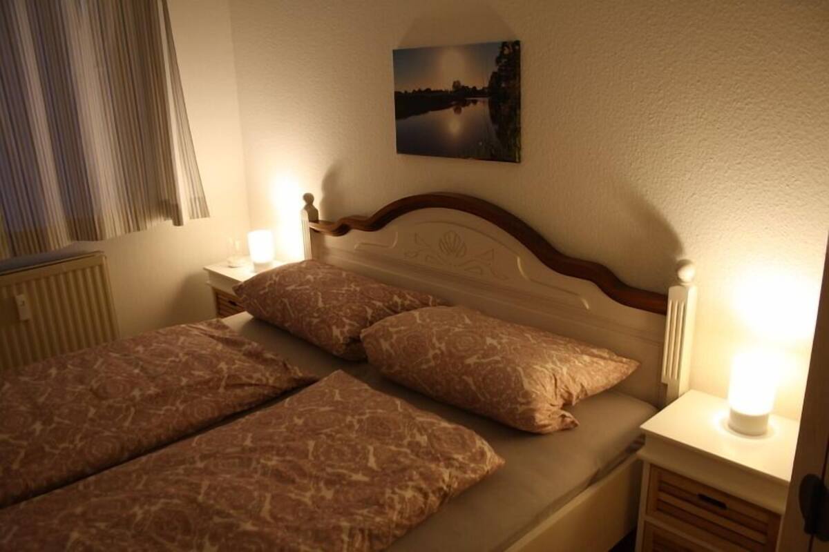 Quarto com cama, duas luminárias de cabeceira, mesa de cabeceira, radiador e um quadro na parede.