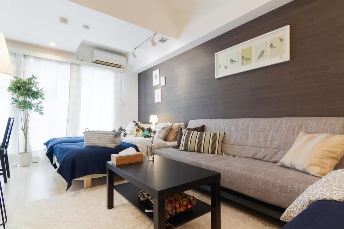 Uma sala de estar moderna com um sofá, uma mesa de centro e uma cama com roupa de cama azul. Há uma planta, um quadro na parede e um aparelho de ar condicionado.