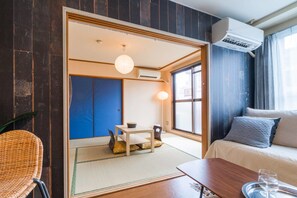Um quarto em estilo japonês tradicional com tatames, uma mesa baixa com uma tigela, um sofá com almofadas e uma porta deslizante para outro espaço.