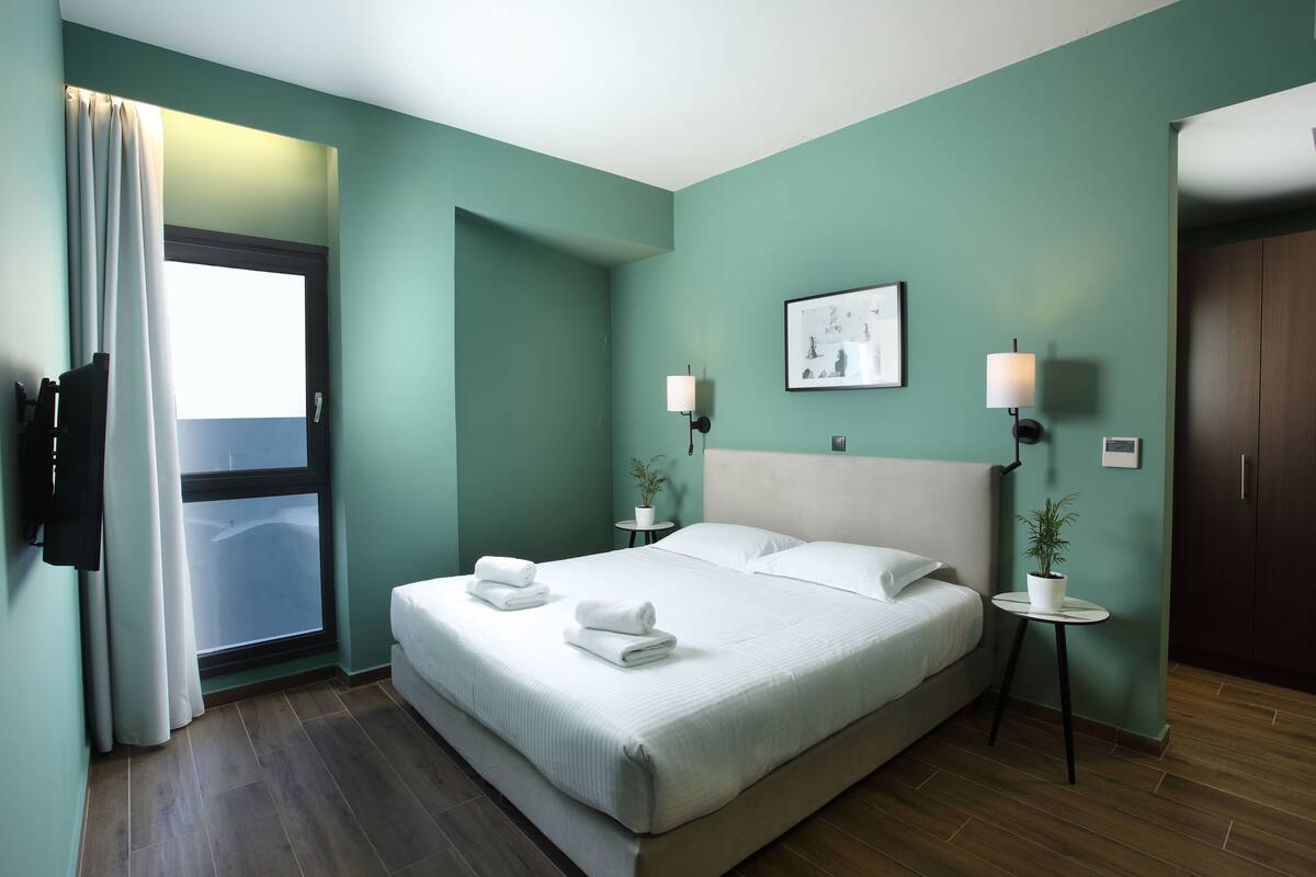 Uma cama bem arrumada com lençóis brancos, uma mesinha de cabeceira com abajur, uma planta em vaso e um quadro na parede.