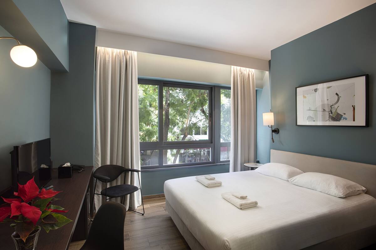 Quarto de hotel com uma cama grande, uma escrivaninha com cadeira, uma janela com cortinas e um candeeiro fixado na parede.