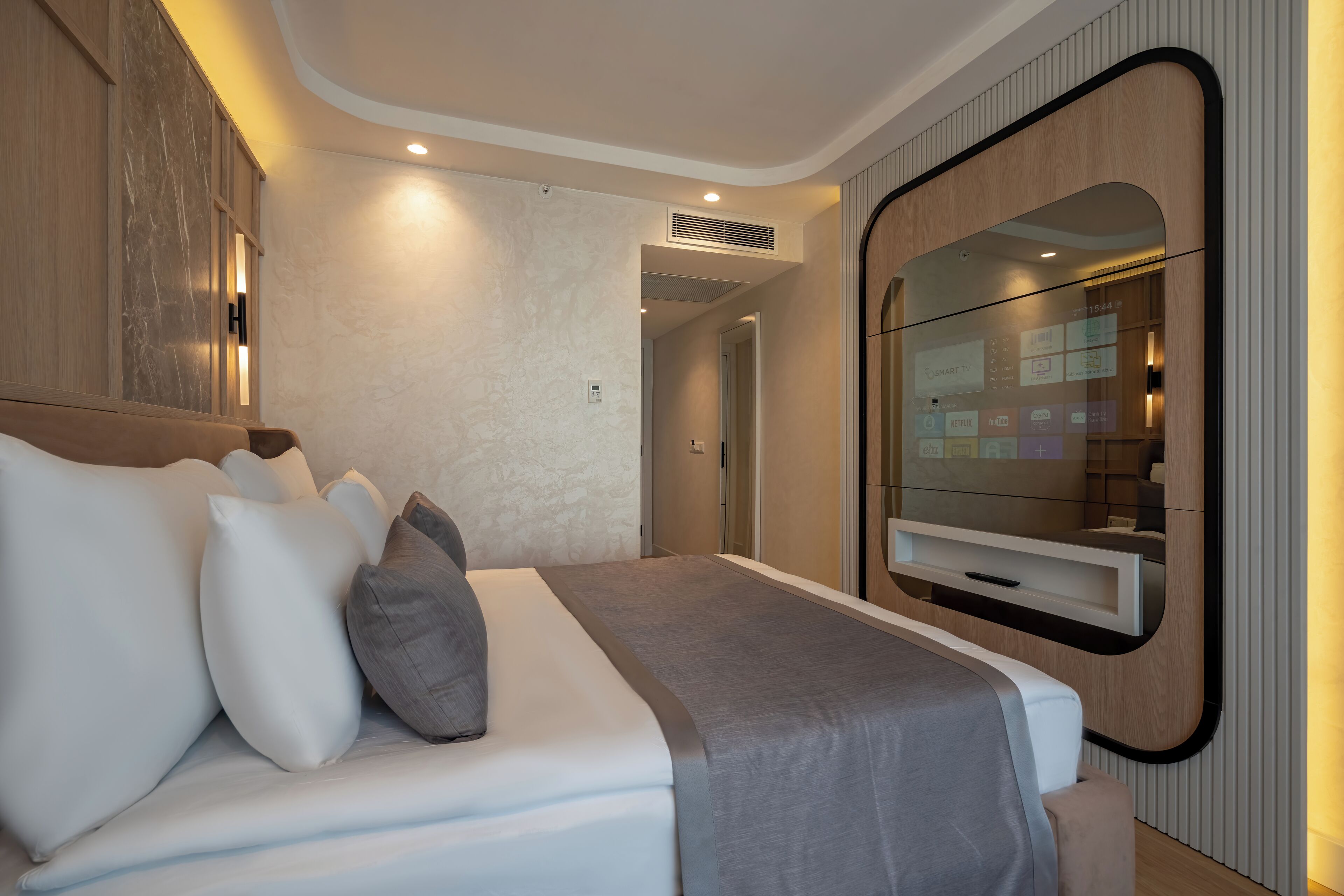 Quarto de hotel moderno com uma cama grande, televisão integrada e espelho com moldura de madeira.