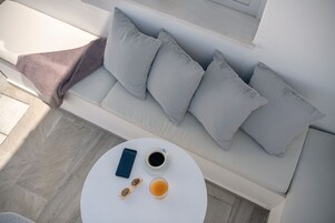 Um sofá branco com almofadas estampadas, uma mesa redonda com um smartphone, uma xícara de café e um copo de chá.