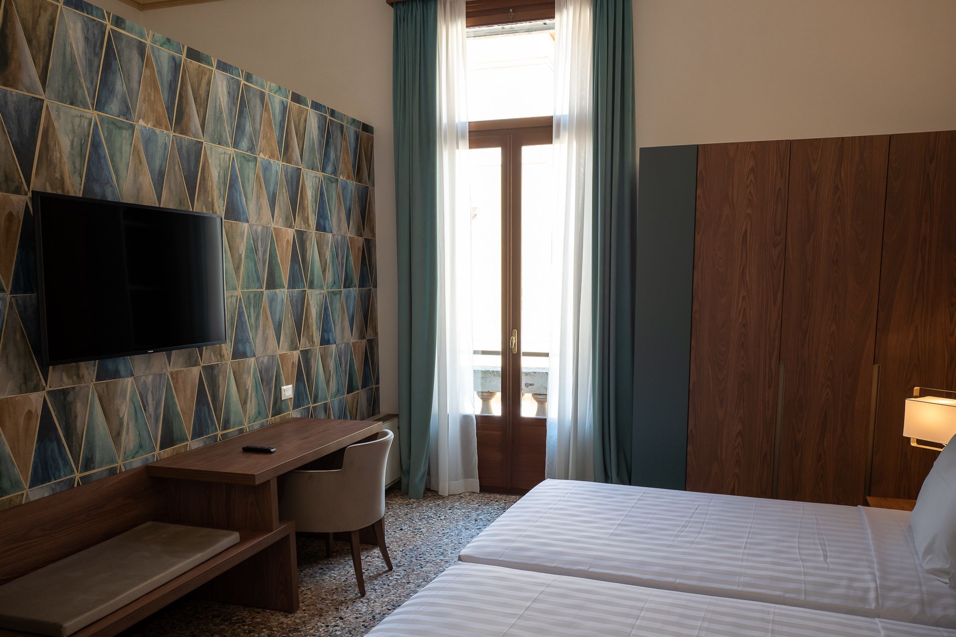 Quarto de hotel com televisão na parede revestida de azulejos, secretária de madeira, cama e janela com cortinas.