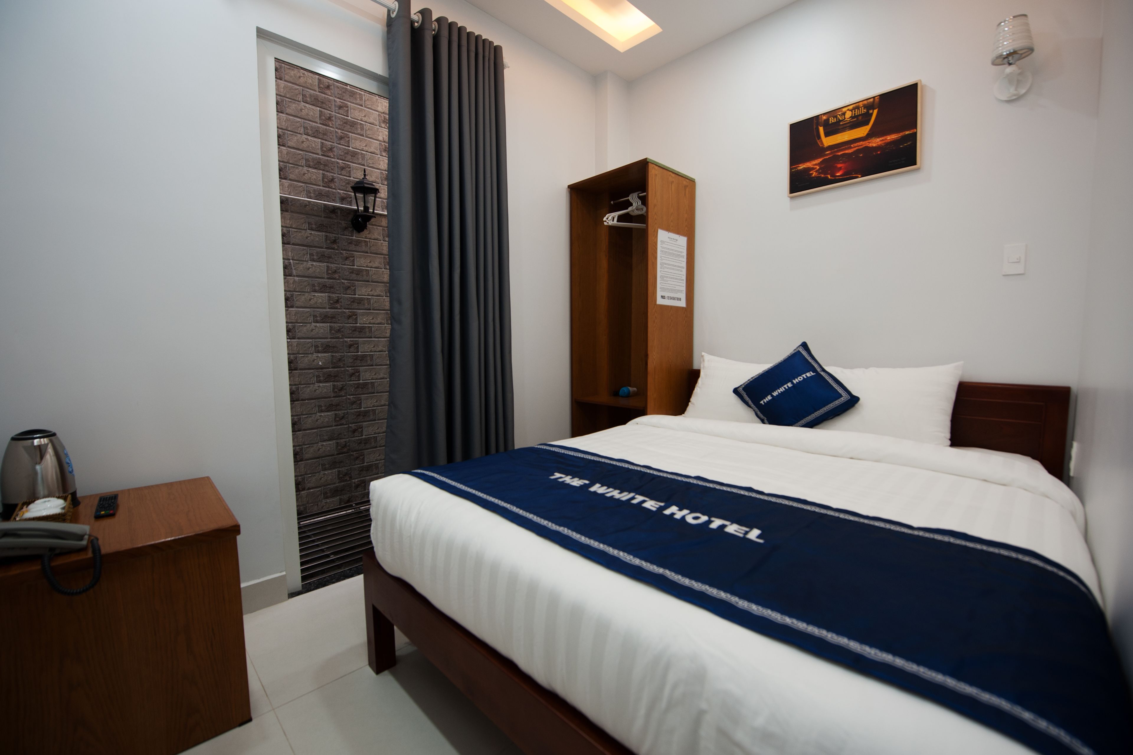 Quarto de hotel com uma cama, um guarda-roupa de madeira, uma escrivaninha, uma cafeteira e uma parede de tijolos.