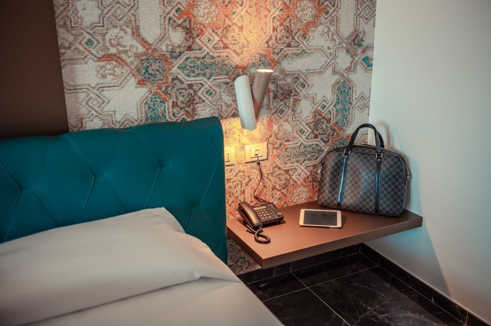 Quarto de hotel com cabeceira verde-água, mesa de cabeceira com abajur, telefone, tablet e bolsa xadrez.