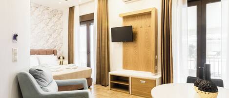 Quarto de hotel moderno com cama, área de estar com mesa, TV fixada na parede e uma janela grande com cortinas.