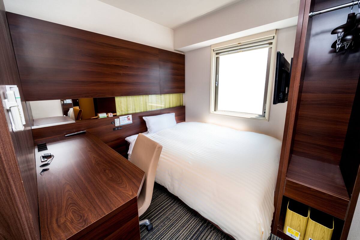 Quarto de hotel com cama, escrivaninha, cadeira, janela e guarda-roupa.
