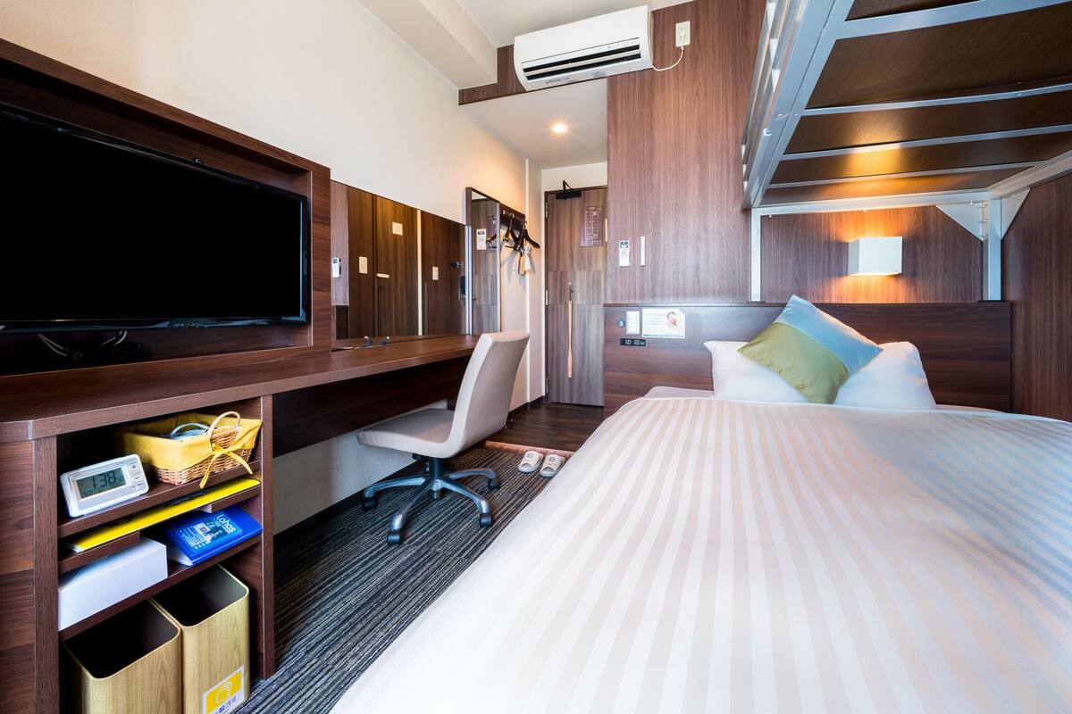 Quarto de hotel com cama, escrivaninha, cadeira, televisão, guarda-roupa e banheiro.