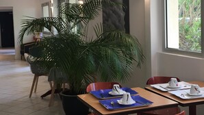 Uma sala de jantar com mesas preparadas para dois, com xícaras e pires brancos, e uma grande planta em vaso ao centro.