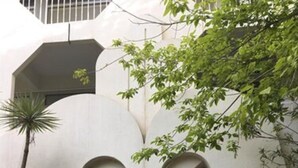 Um edifício com design arquitetônico moderno, destacando uma entrada central com portas de vidro e piso ladrilhado. Vasos com plantas e áreas de estar ao ar livre são visíveis.