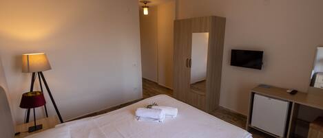 Quarto de hotel com cama bem arrumada, escrivaninha com televisão e pequena geladeira.