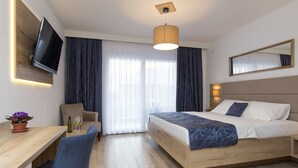 Quarto de hotel com cama, escrivaninha, cadeira, televisão e janela com cortinas.