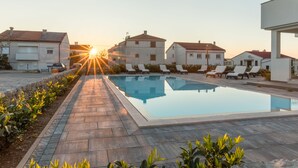 Piscina com espreguiçadeiras, um caminho pavimentado e edifícios ao fundo durante o pôr do sol.
